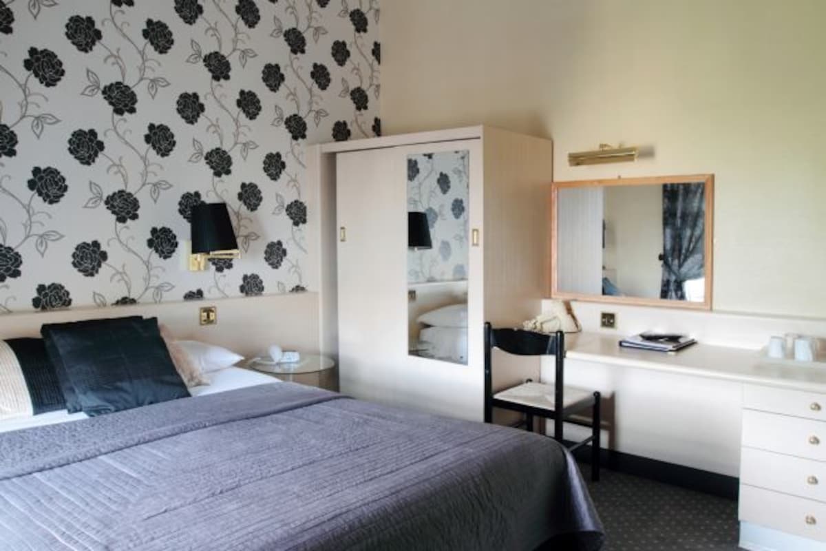 Quarto de hotel com cama, escrivaninha, cadeira, guarda-roupa e espelho.
