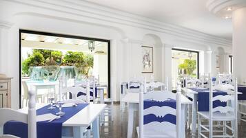 Restaurante com mesas e cadeiras brancas, toalhas de mesa azuis e vista para um jardim pelas portas abertas.