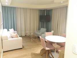 Uma sala de estar moderna com um sofá branco, uma mesa redonda com cadeiras cor-de-rosa, uma mesa de centro de vidro e uma televisão de tela plana fixada na parede.