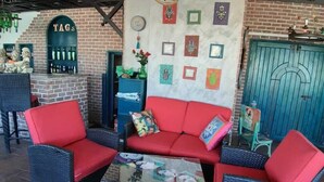 Uma área de estar coberta ao ar livre com móveis de vime e almofadas vermelhas, uma parede de tijolos com quadros e uma porta azul.