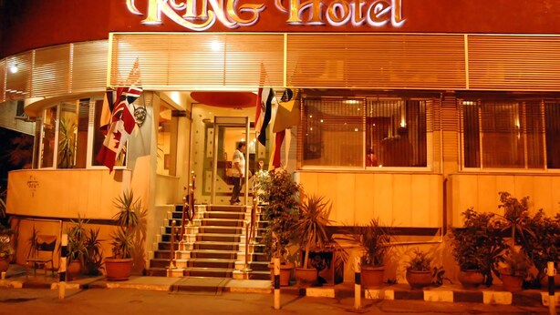 Exterior do HOTEL KING com uma placa visível, uma escadaria que leva à entrada e plantas em vasos na área da entrada.