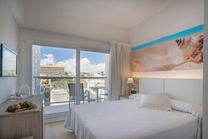 Quarto de hotel com uma cama, uma escrivaninha com uma bandeja de frutas, uma cadeira e vista da cidade por grandes janelas.