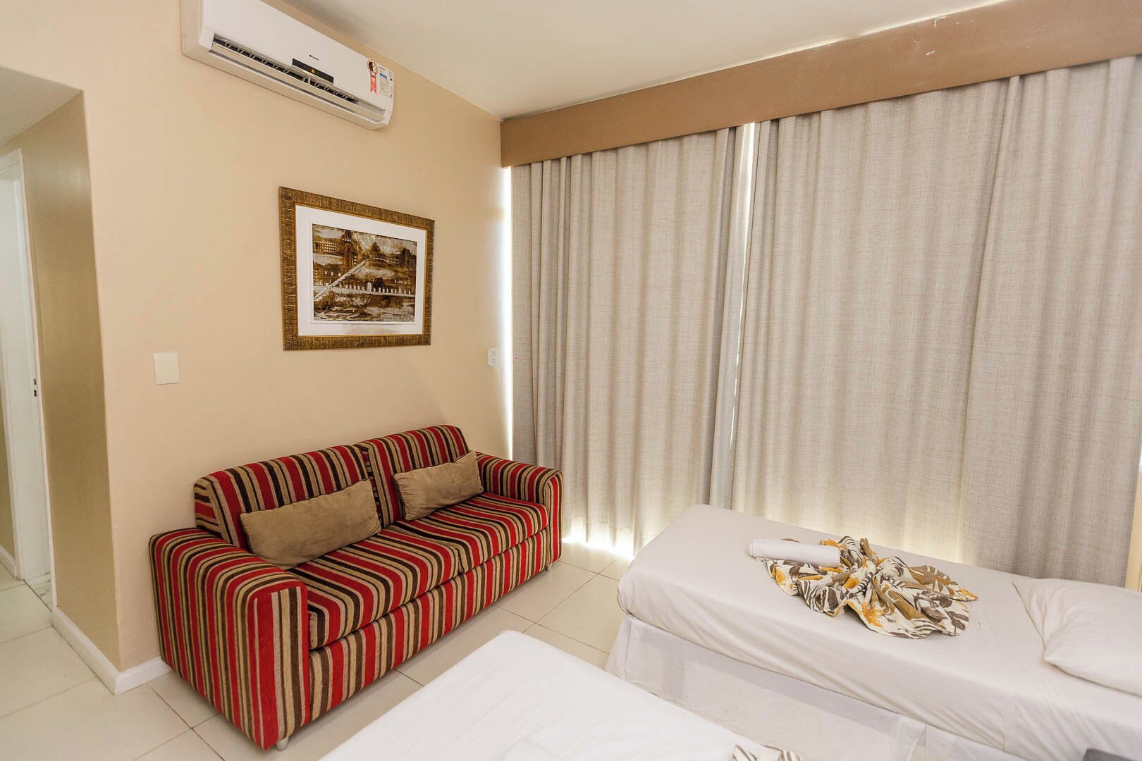 Quarto com sofá listrado, cama com manta estampada, quadro pendurado na parede e janela com cortinas.