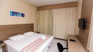Quarto de hotel com cama, escrivaninha, cadeira, televisão e janela com cortinas.