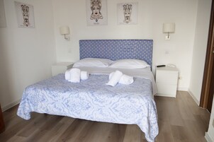 Uma cama bem arrumada com colcha estampada, duas mesas de cabeceira e luminárias fixadas na parede.