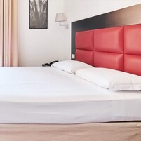Uma cama bem arrumada com lençóis brancos, cabeceira estofada vermelha e uma luminária fixada na parede.