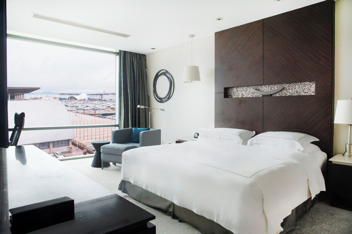 Quarto de hotel moderno com uma cama grande, escrivaninha, cadeira e vista para a paisagem urbana.