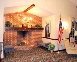 Quarto aconchegante com lareira de tijolos, um candelabro e uma bandeira. Há duas poltronas e uma mesa com livros.