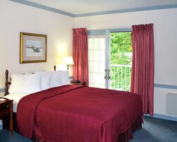 Quarto de hotel com uma cama grande, uma mesa de cabeceira, uma luminária, um quadro na parede e vista para o verde pela janela.