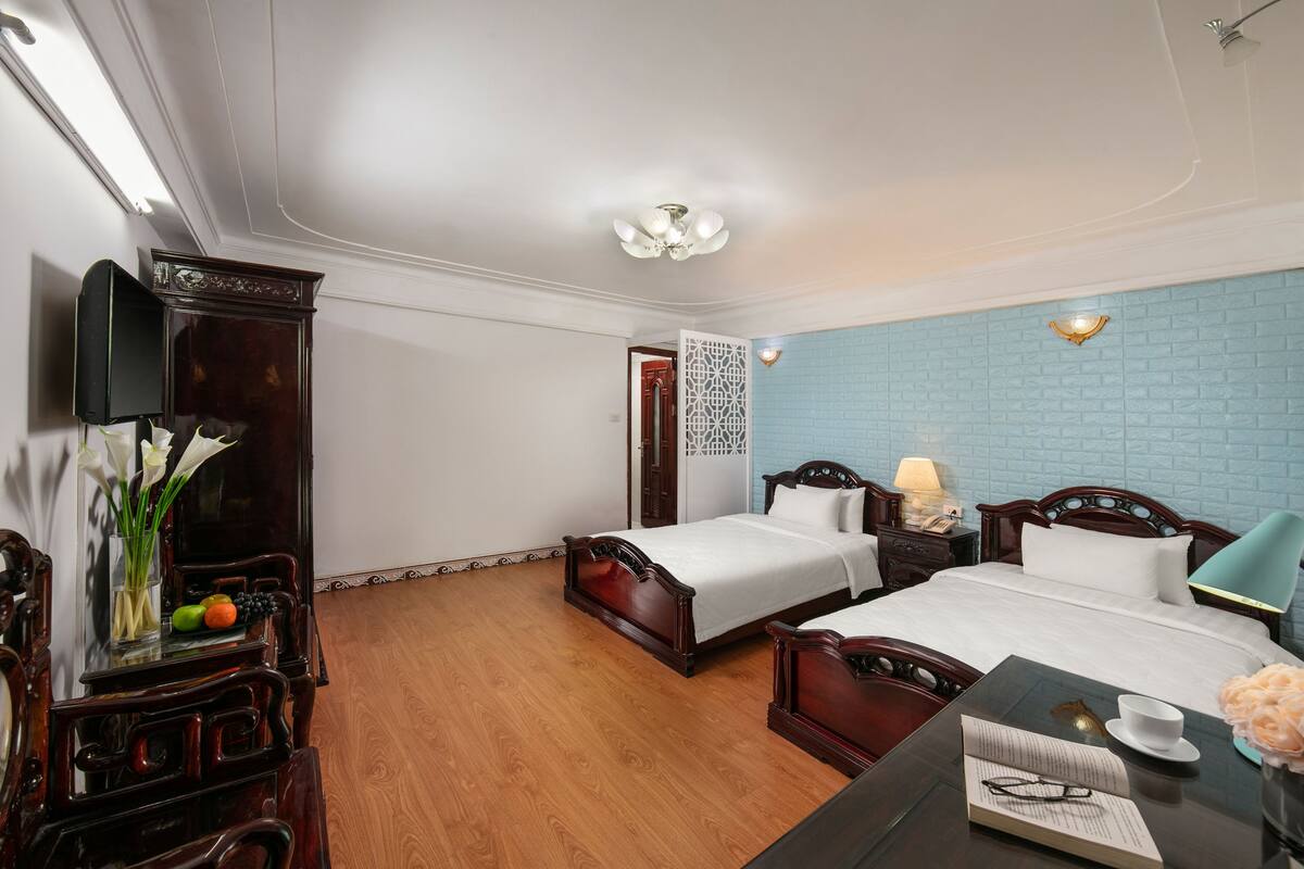 Quarto de hotel com duas camas, uma cômoda de madeira, uma mesa com abajur e uma parede de tijolos azuis.