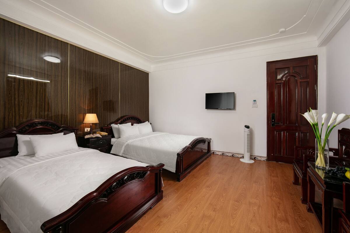 Quarto de hotel com duas camas de madeira, uma TV na parede, um aquecedor portátil e uma porta de madeira.
