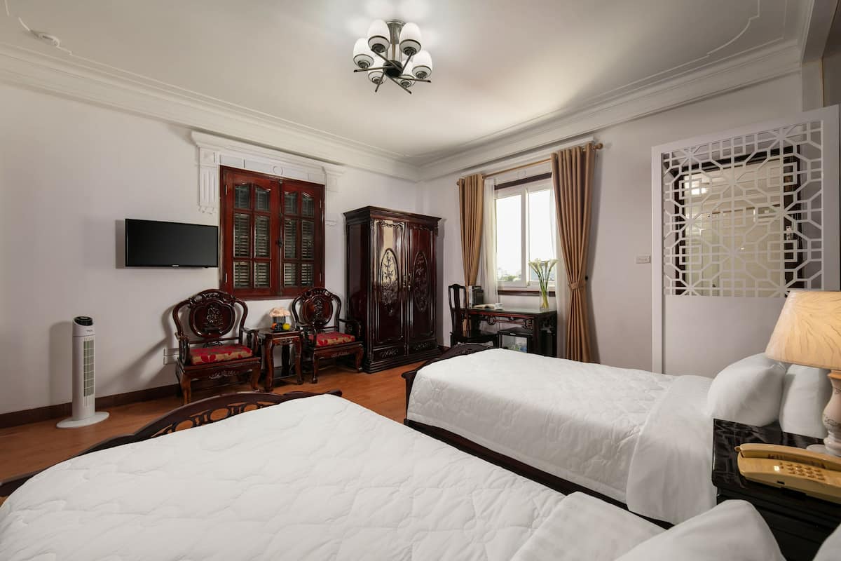 Quarto de hotel com duas camas, televisão, guarda-roupa, secretária com candeeiro e janela com cortinas.
