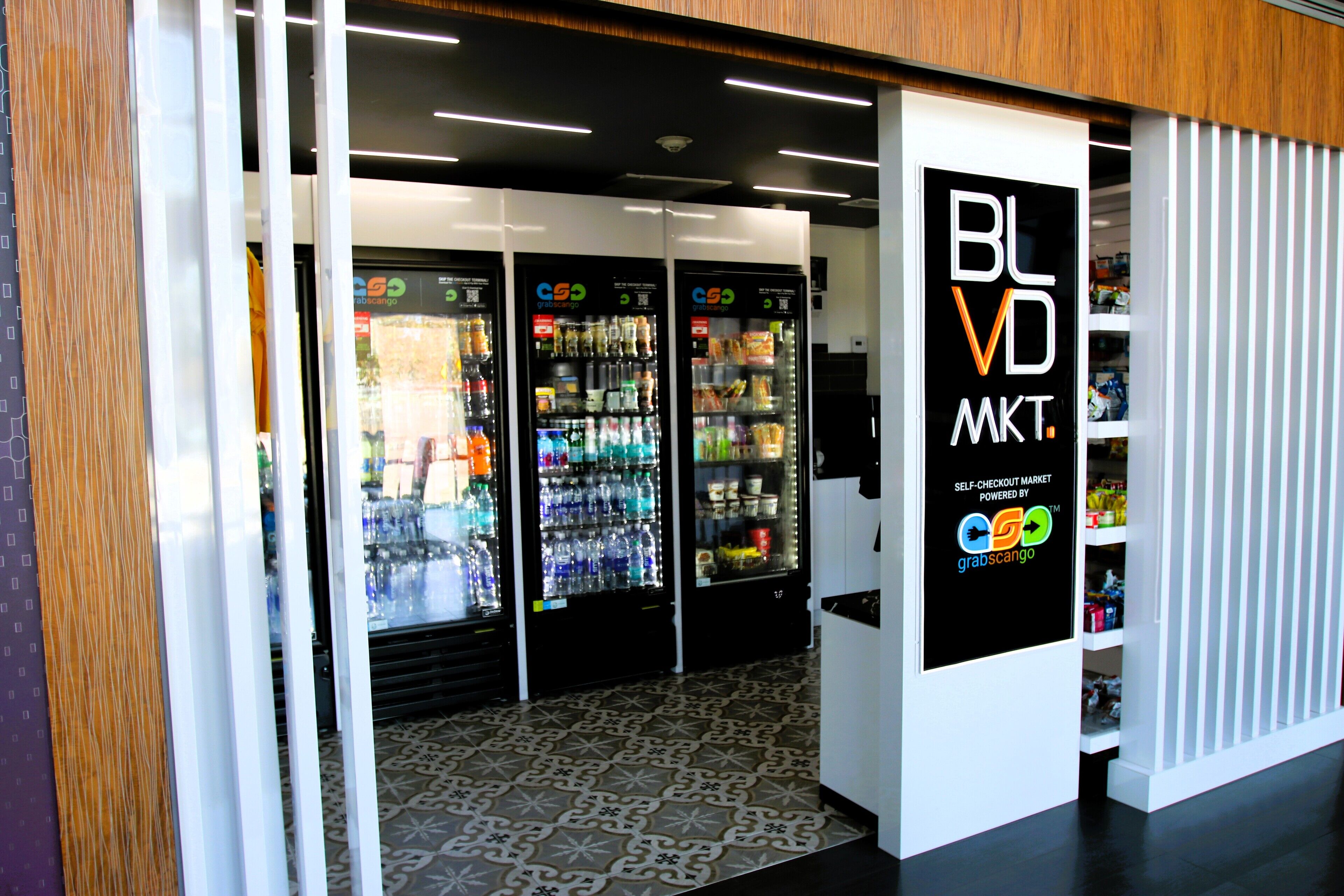 Uma área de autoatendimento com três máquinas de venda automática de bebidas e uma placa com a inscrição 'BLVD MKT. powered by grabscango'.