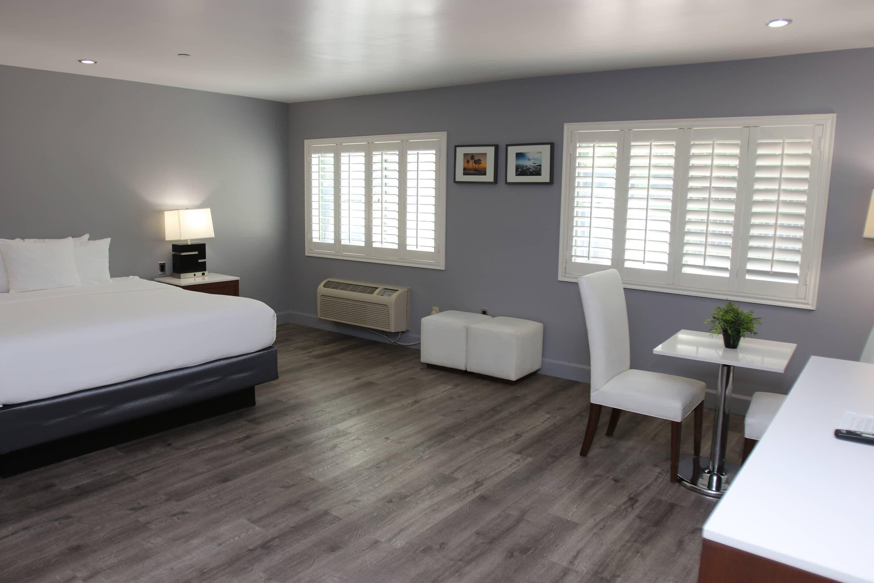 Quarto de hotel com cama, mesa de cabeceira, luminária, ar condicionado, dois pufes brancos, uma pequena mesa com uma planta e duas janelas com persianas.