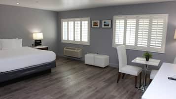Quarto de hotel com cama, mesa de cabeceira, luminária, ar condicionado, dois pufes brancos, uma pequena mesa com uma planta e duas janelas com persianas.