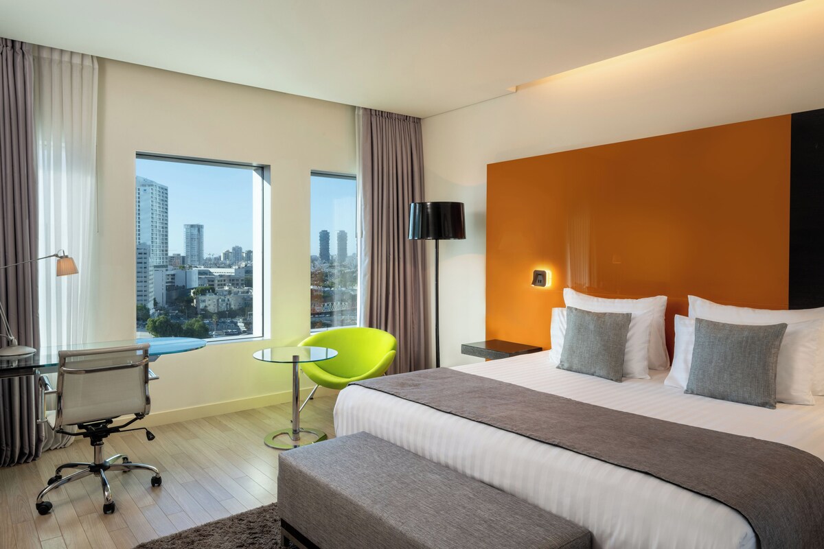 Quarto de hotel moderno com uma cama grande, uma escrivaninha, uma poltrona verde vibrante e vista para a cidade através de amplas janelas.
