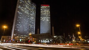 Dois arranha-céus imponentes com janelas iluminadas e sinalização, uma estrutura curva em primeiro plano e rastros de luz deixados por veículos em movimento.