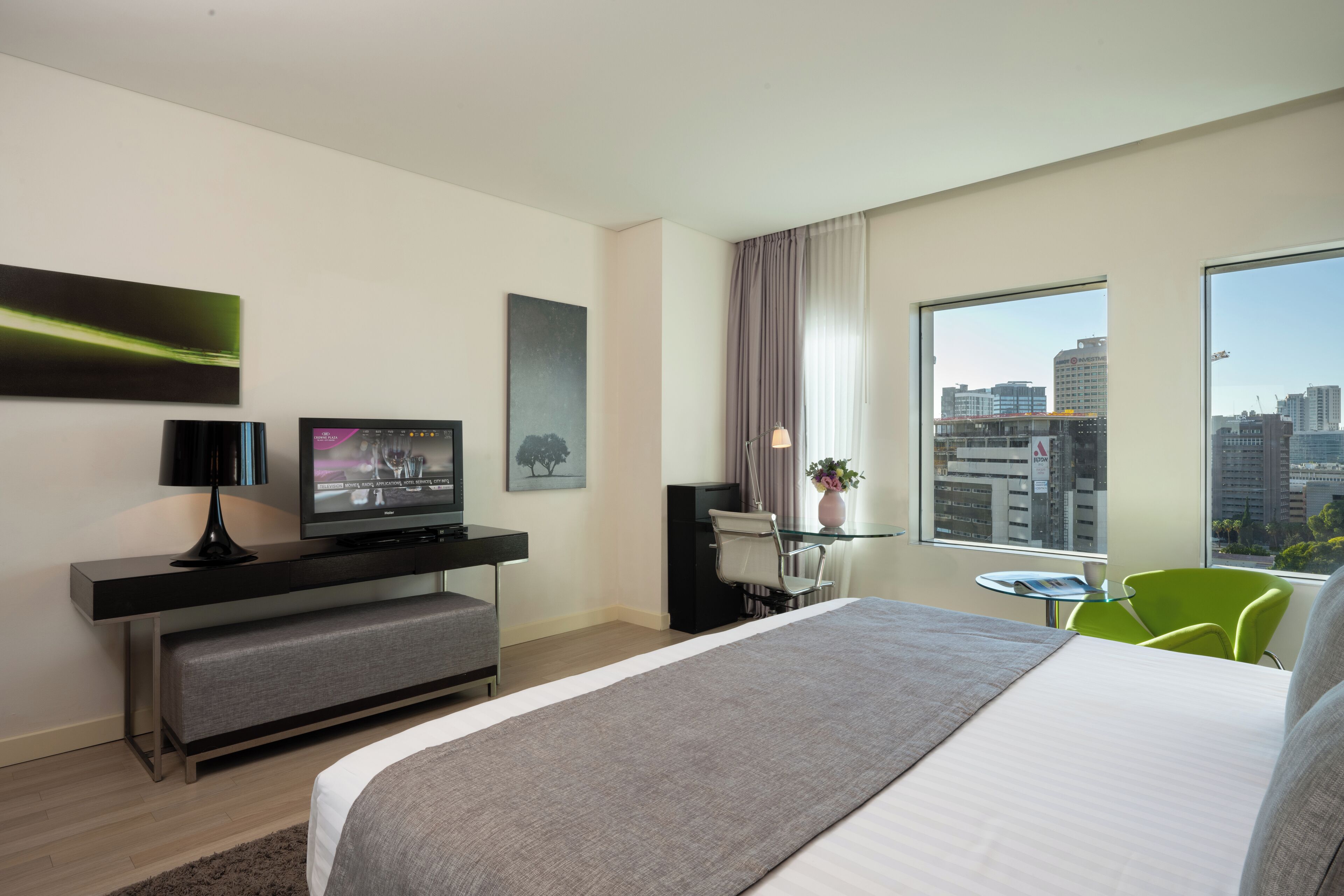 Quarto de hotel moderno com uma cama grande, uma escrivaninha com televisão de tela plana, uma cadeira, uma mesinha de cabeceira com abajur e vista para a cidade através de grandes janelas.