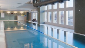 Piscina interior com paredes de vidro transparente, posto de salva-vidas e vista para a cidade através de amplas janelas.
