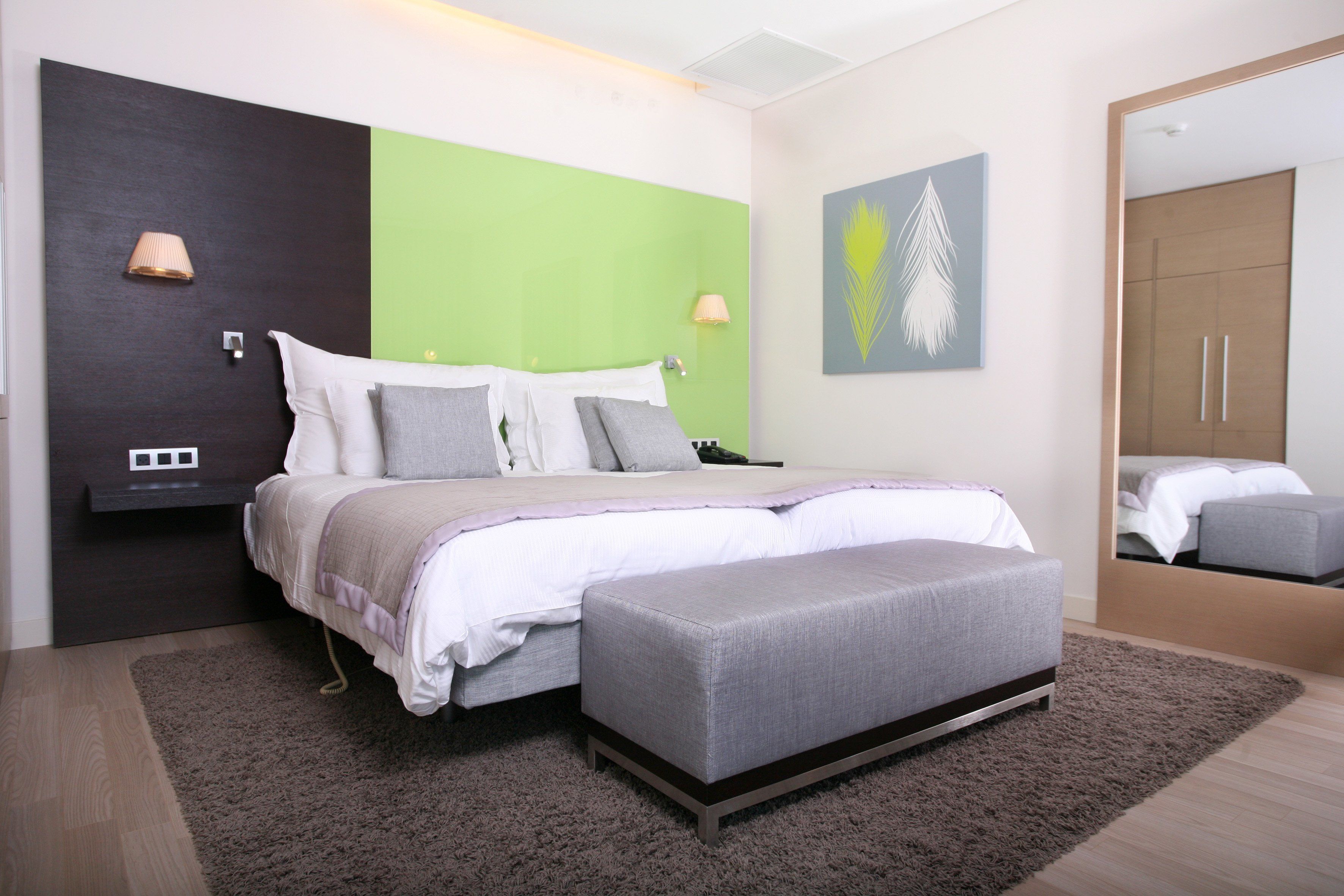 Quarto moderno com cama, abajures, um banco cinza e uma parede verde destacada.
