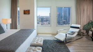 Quarto de hotel moderno com uma cama grande, um espaço de estar com uma poltrona, vista para a cidade e uma planta em vaso.