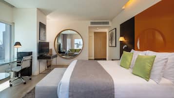 Quarto de hotel moderno com uma cama grande, uma escrivaninha com computador, televisão e uma janela ampla com vista para a cidade.