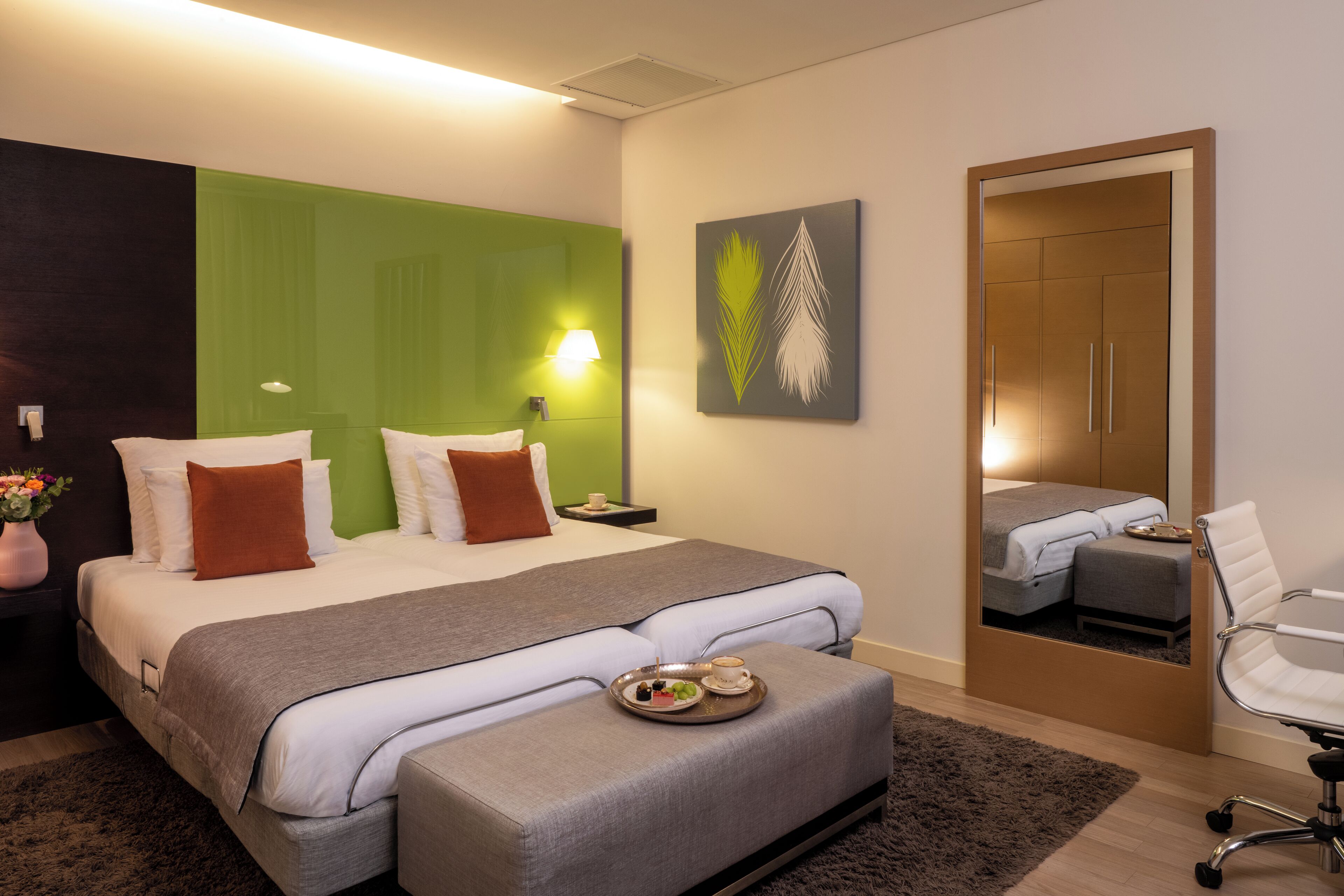 Quarto de hotel moderno com uma cama grande, uma mesa de cabeceira com abajur, uma pequena mesa com bandeja de comida e uma xícara, e um armário com espelho.