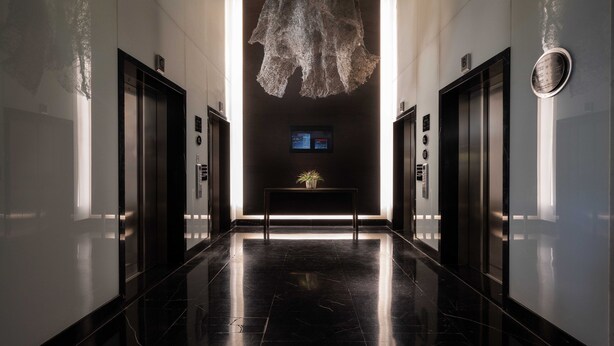Corredor moderno com pisos de mármore preto, dois elevadores, balcão de recepção com planta e um grande candelabro.