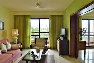 Uma sala de estar com sofá, mesa de centro com objetos decorativos, televisão e varanda com cortinas.