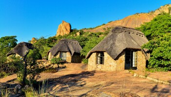 Huts com telhados de palha e paredes de pedra, inseridas numa paisagem natural com uma grande formação rochosa ao fundo.