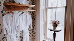 Quarto de canto com papel de parede floral, piso de madeira, roupão branco pendurado, mesa redonda pequena com um vaso e uma grande janela em arco.