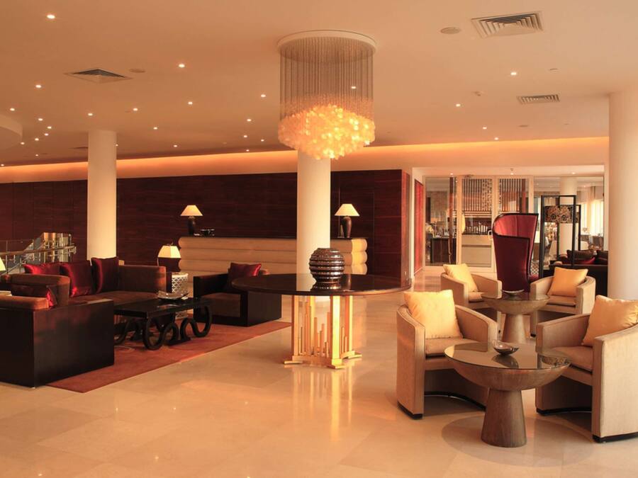 Um hall amplo com design moderno, apresentando uma área central de estar com sofás e poltronas, um lustre grande e um balcão de recepção.