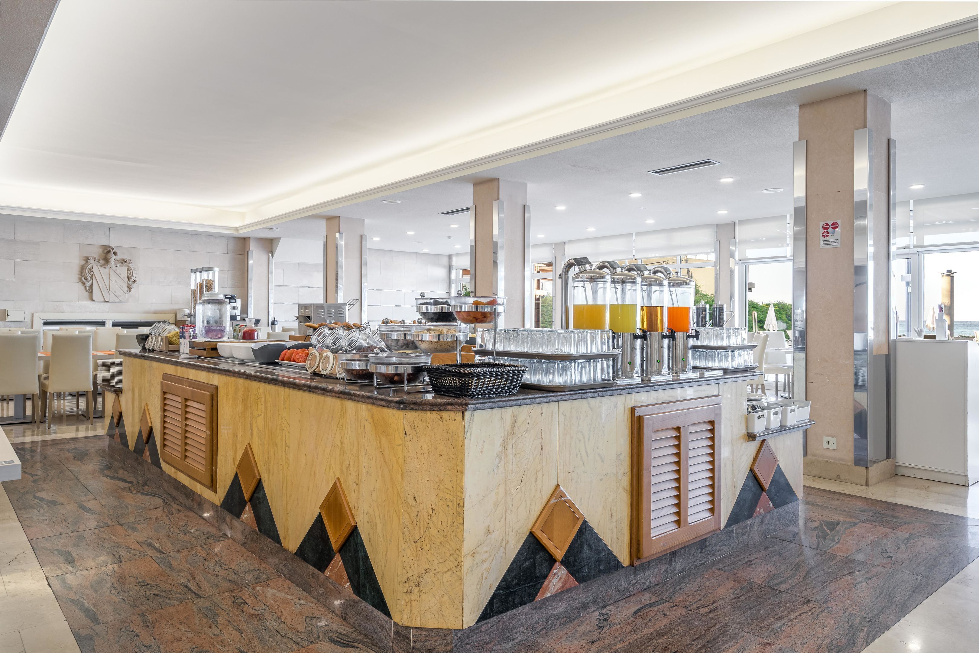 Um balcão de café da manhã moderno com bancada de mármore, diversos distribuidores de comida e bebida, e uma área de estar com cadeiras.