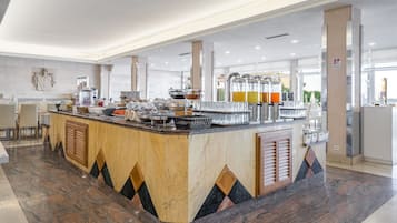 Um balcão de café da manhã moderno com bancada de mármore, diversos distribuidores de comida e bebida, e uma área de estar com cadeiras.