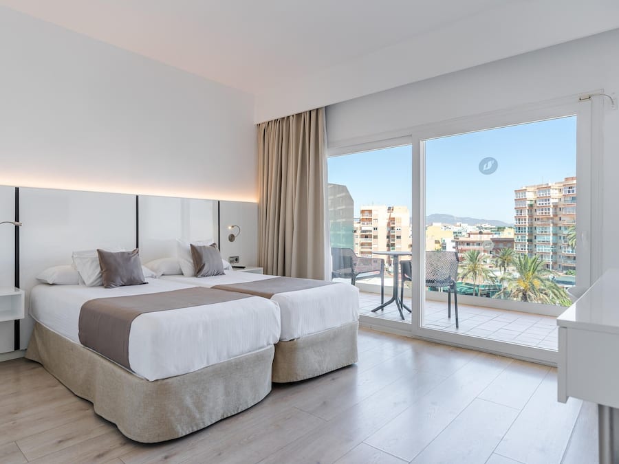 Quarto de hotel moderno com uma cama grande, mesa de cabeceira com telefone e vista para a cidade através de amplas janelas.