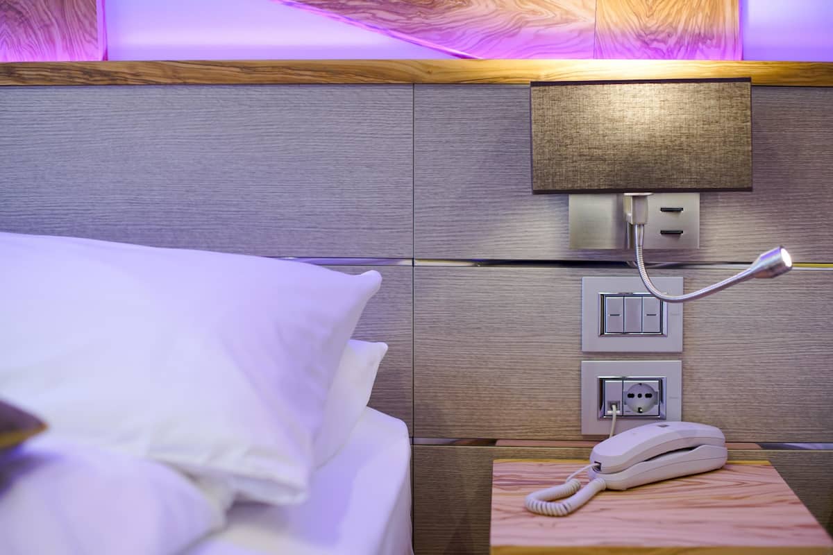Quarto de hotel com cama arrumada, mesa de cabeceira com telefone e tomada na parede.