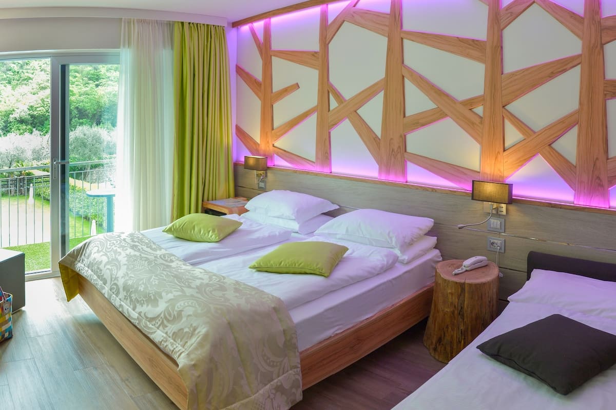 Quarto de hotel com uma cama grande, duas mesas de cabeceira, uma cabeceira de madeira e uma varanda com vista para o verde.