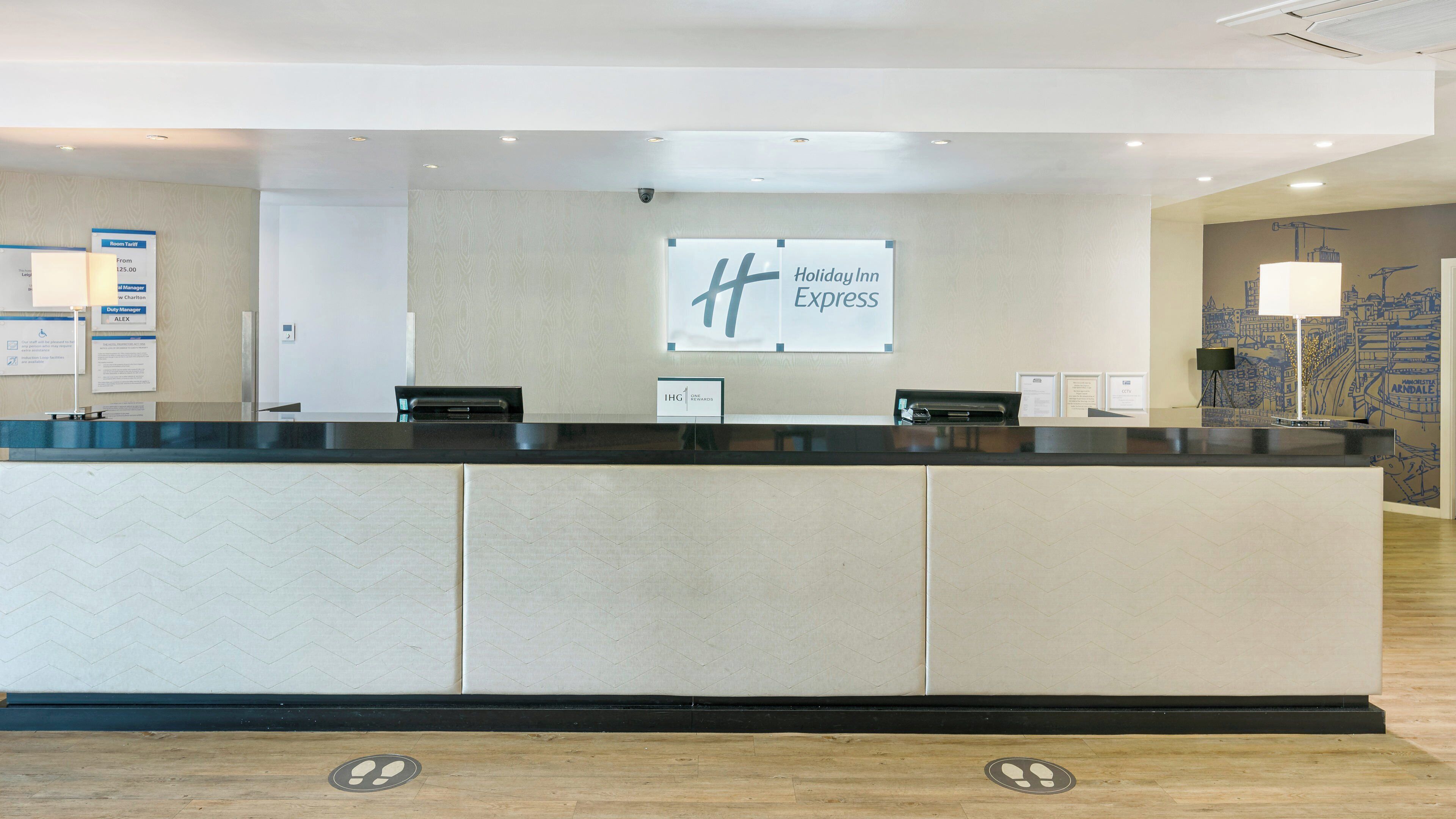 Bancada de recepção de hotel com uma placa grande da Holiday Inn Express, uma tela digital e uma área de recepção com parede estampada e um mural com vista para a cidade.
