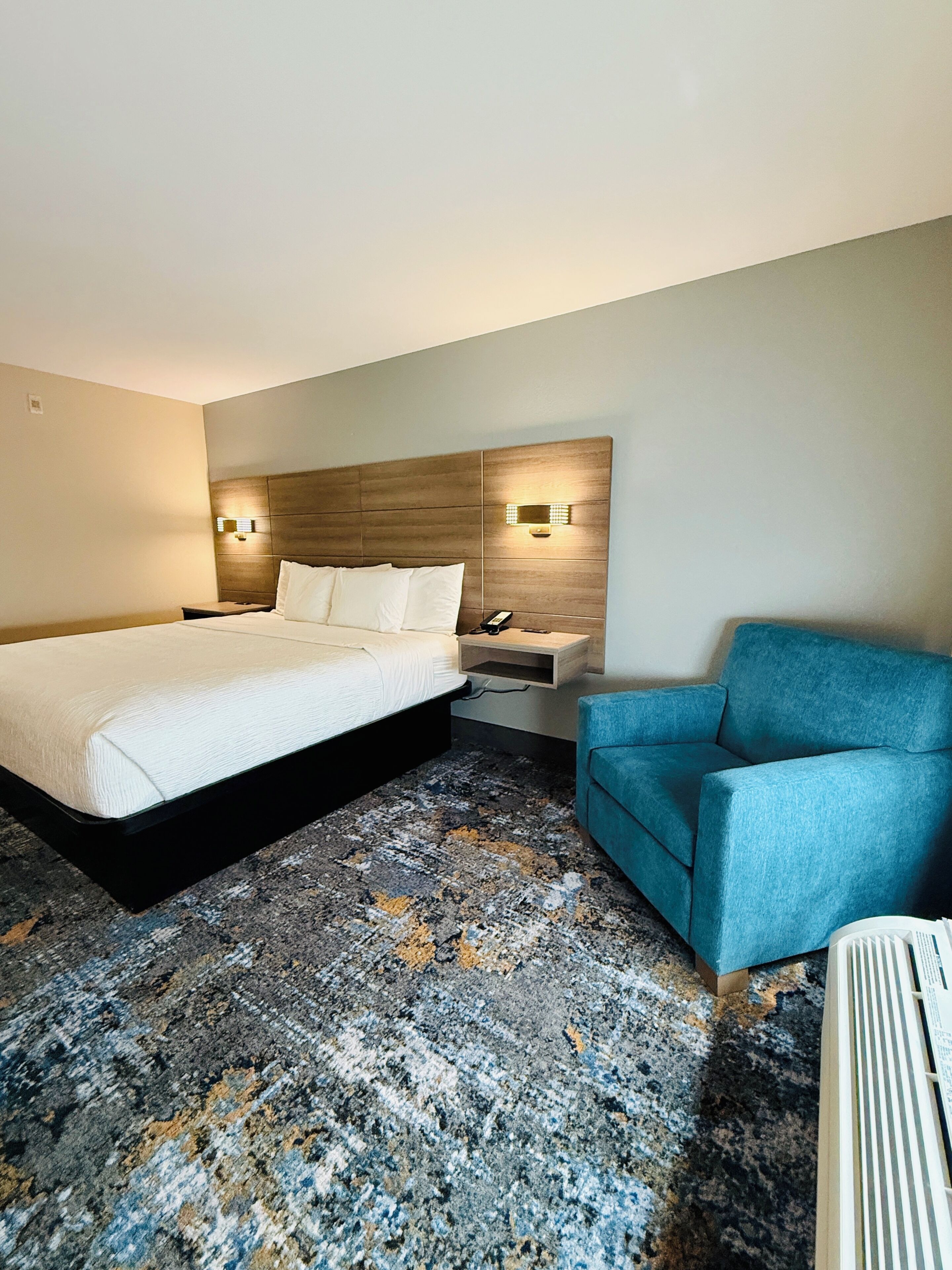 Quarto de hotel com cama, mesas de cabeceira, uma poltrona azul e um tapete estampado.