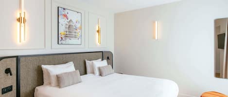 Quarto de hotel com uma cama grande, duas luminárias fixadas na parede, um quadro emoldurado e um pequeno banquinho.