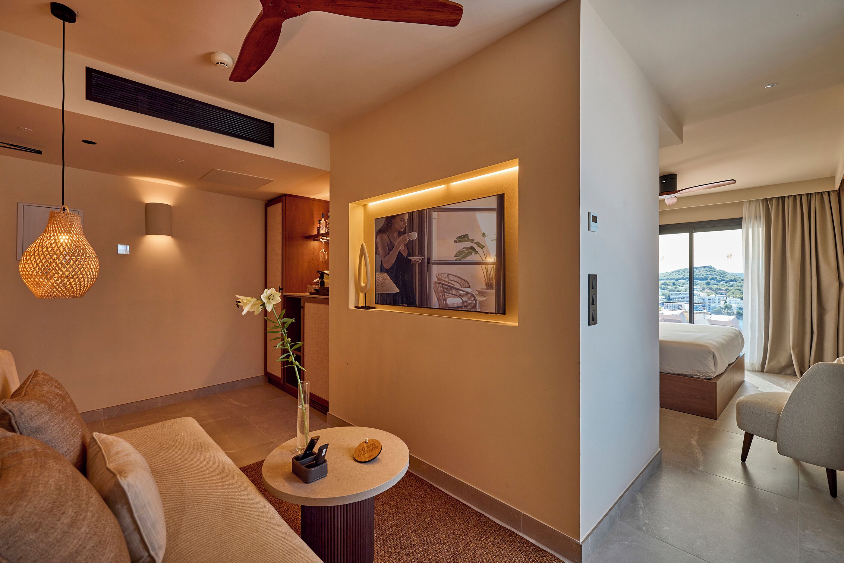 Quarto de hotel moderno com sofá, mesa redonda com vaso e objeto decorativo, pintura acima da cama e vista da cidade pela janela.