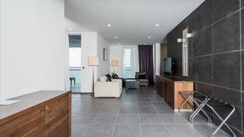 Quarto de hotel moderno com cômoda de madeira, um sofá, televisão e área de refeições com um banquinho.