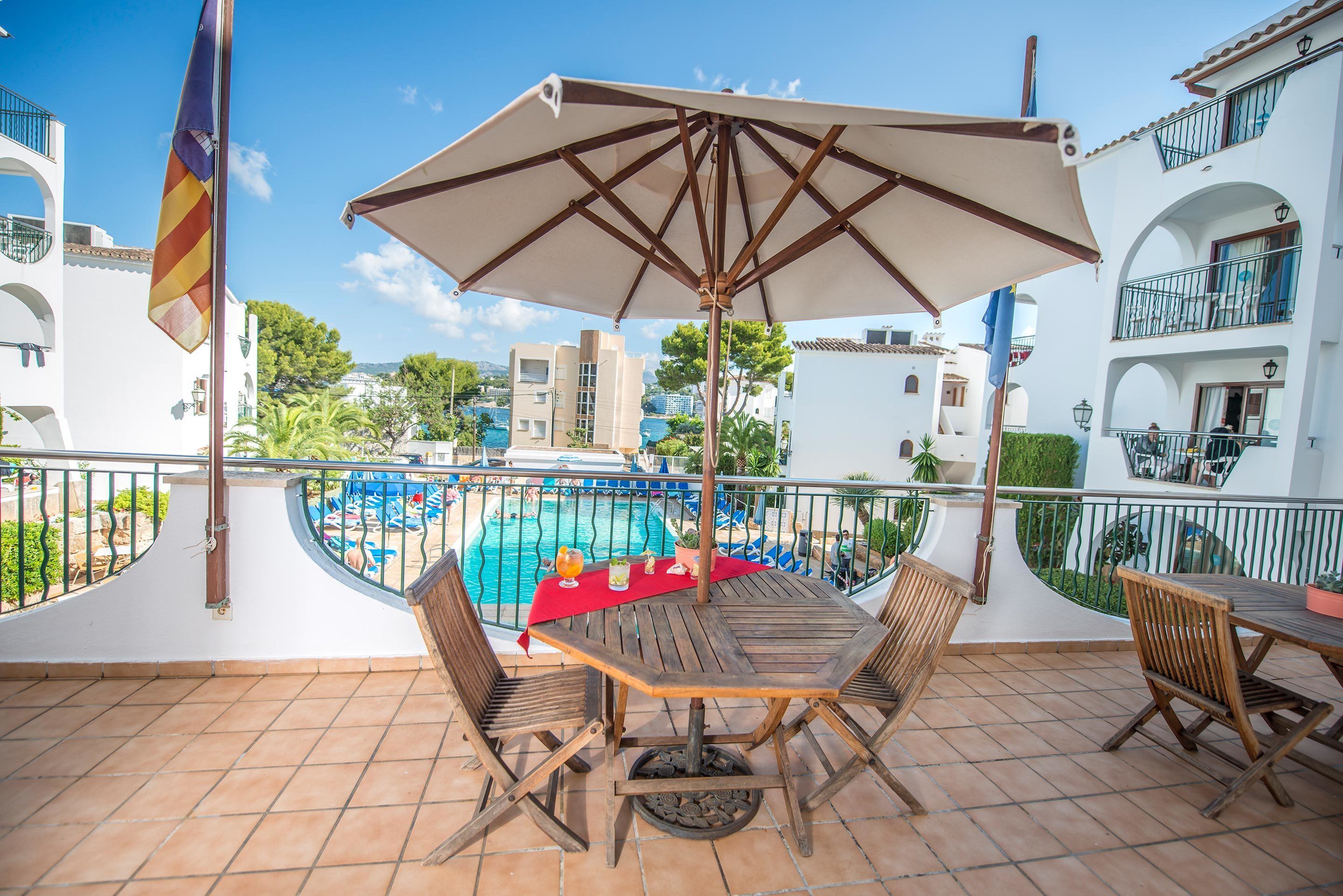 Um terraço com mesa de madeira, cadeiras e um grande guarda-sol. Uma bandeira é visível e uma piscina ao fundo.