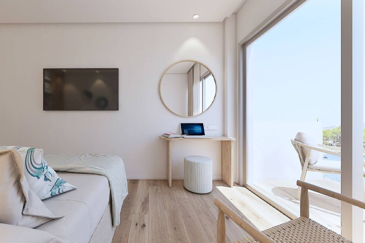 Quarto de hotel moderno com uma cama, uma escrivaninha com um laptop, um espelho redondo e vista para o exterior.