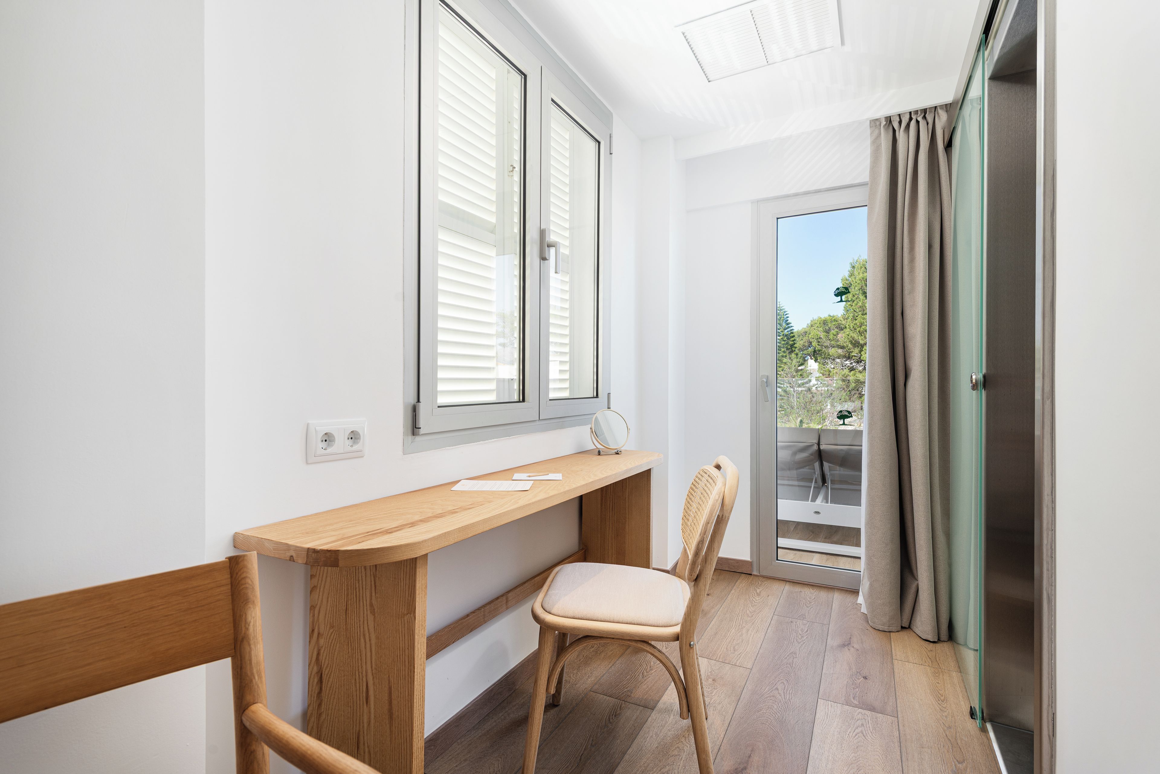 Quarto com escrivaninha de madeira, cadeira, janela com persianas, porta e vista para o exterior.