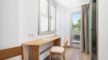 Quarto com escrivaninha de madeira, cadeira, janela com persianas, porta e vista para o exterior.