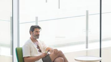 Um homem sentado numa cadeira verde em uma sala moderna com janelas grandes, uma mesa redonda e piso de mármore.