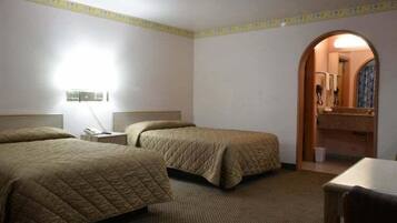 Quarto de hotel com duas camas, uma escrivaninha, uma cadeira, uma mesa de cabeceira, um abajur e um banheiro visível por um arco.