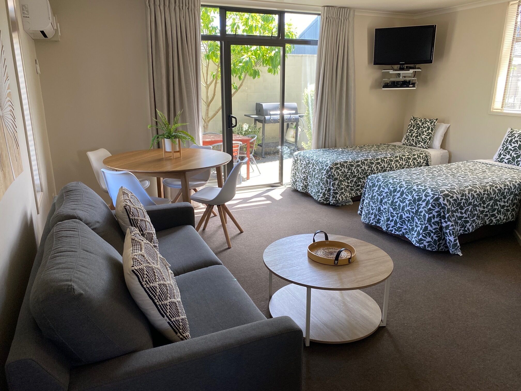 Quarto de hotel moderno com sofá, duas camas de solteiro, uma mesa de jantar com cadeiras, televisão e varanda com churrasqueira.