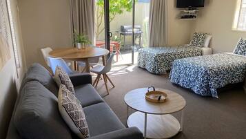 Quarto de hotel moderno com sofá, duas camas de solteiro, uma mesa de jantar com cadeiras, televisão e varanda com churrasqueira.